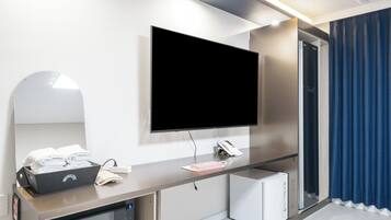Quarto de hotel com televisão de tela plana na parede, uma pequena escrivaninha com telefone, um mini-refrigerador e uma caixa de artigos de higiene pessoal.