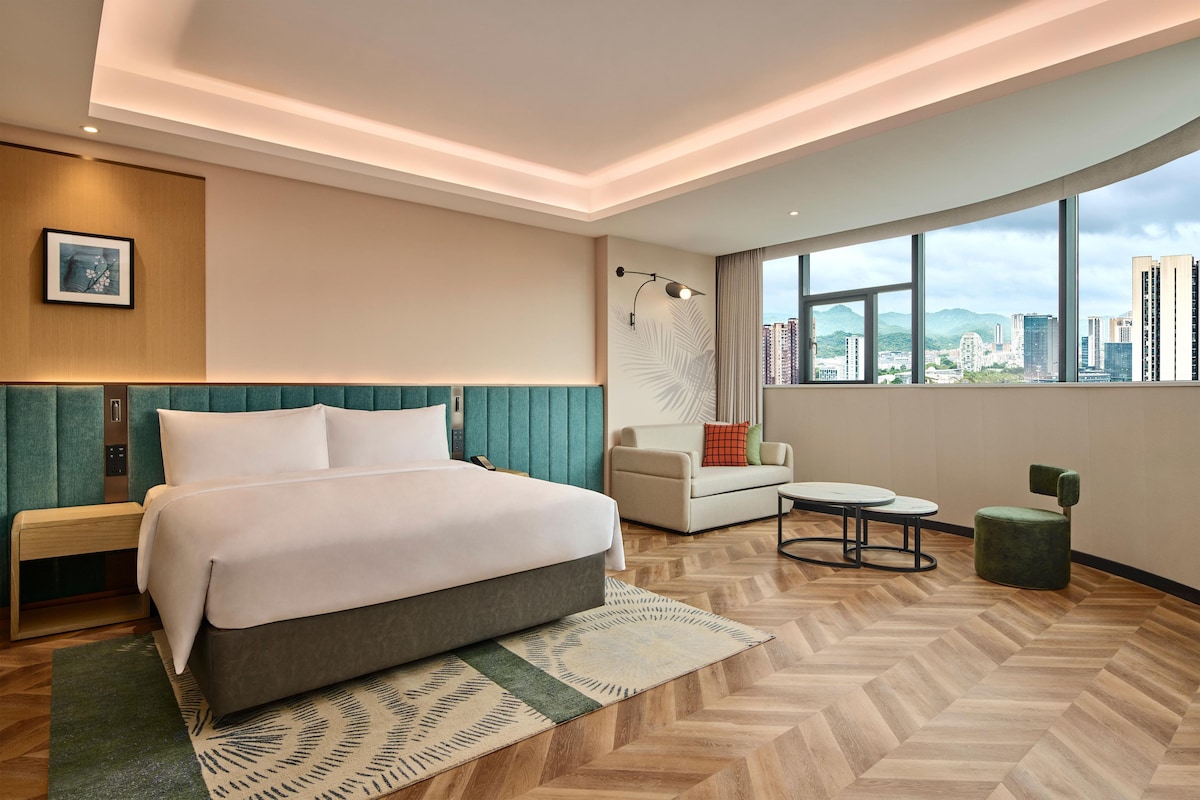 Quarto de hotel com cama, sofá, mesa pequena, banquinho verde e uma grande janela com vista para a cidade.