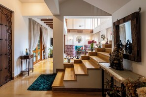 Um interior espaçoso com uma escadaria que leva a um andar superior, uma porta de madeira com detalhes entalhados e diversos objetos decorativos, incluindo estátuas e obras de arte nas paredes.
