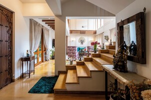 Um interior espaçoso com uma escadaria que leva a um andar superior, uma porta de madeira com detalhes entalhados e diversos objetos decorativos, incluindo estátuas e obras de arte nas paredes.