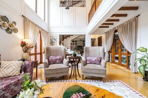 Uma sala de estar ampla com duas poltronas, um sofá, uma mesa de centro e um tapete decorativo. Uma escada é visível ao fundo, dando acesso a um andar superior.