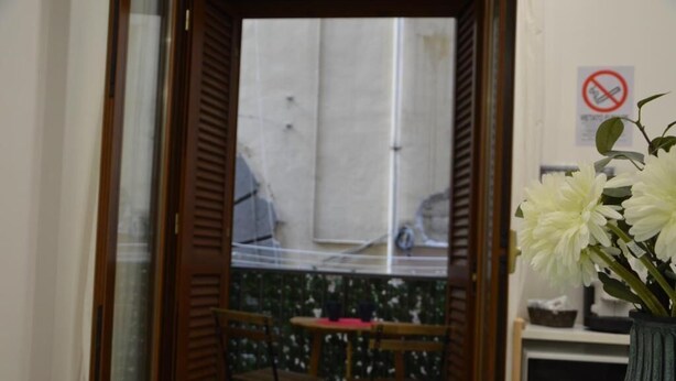 Um quarto com porta de madeira, janela com persianas, uma mesa pequena com cadeiras e um vaso com flores.