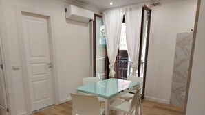 Uma área de refeições com mesa de vidro e cadeiras brancas, uma porta branca e uma janela com cortinas.