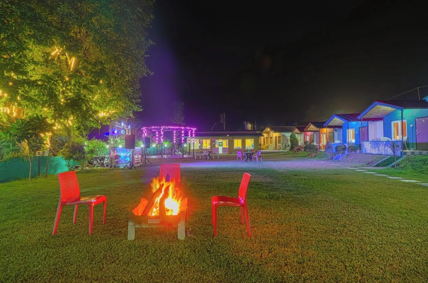 Área externa noturna com lareira e cadeiras, cercada por edifícios com fachadas iluminadas.