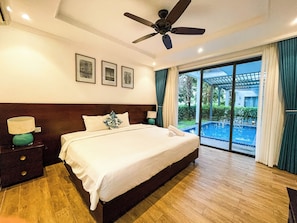 Quarto de hotel com uma cama grande, um ventilador de teto, mesa de cabeceira com abajur e vista para uma piscina pela porta de vidro.