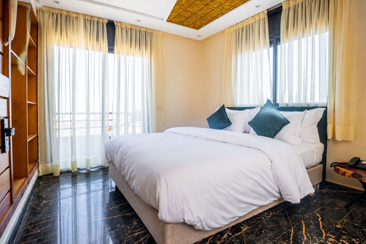 Quarto de hotel com cama, guarda-roupa de madeira, mesa de cabeceira com telefone e janela com cortinas.