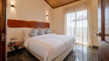 Quarto de hotel com uma cama grande, cabeceira de madeira, mesa de cabeceira com telefone e janela com cortinas translúcidas.