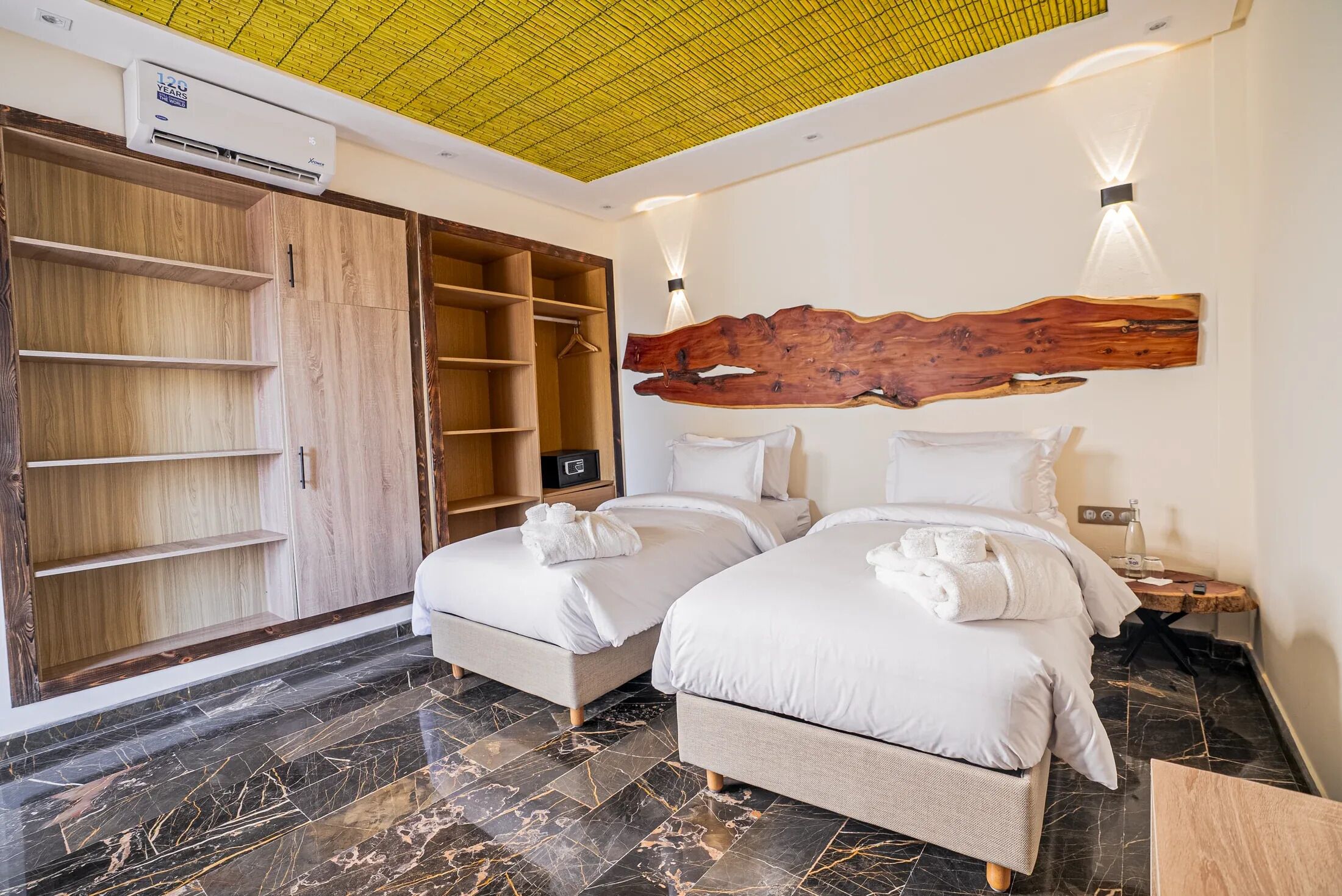 Quarto de hotel com duas camas, um guarda-roupa, cabeceira de madeira, piso de mármore e teto com padrão quadriculado amarelo.