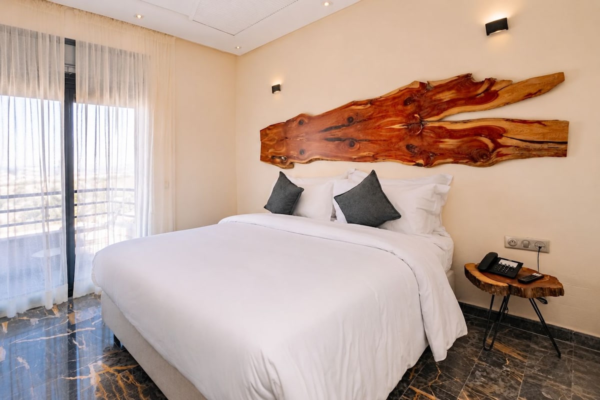 Quarto de hotel com uma cama grande, cabeceira de madeira, mesa de cabeceira com telefone e janela com cortinas finas.