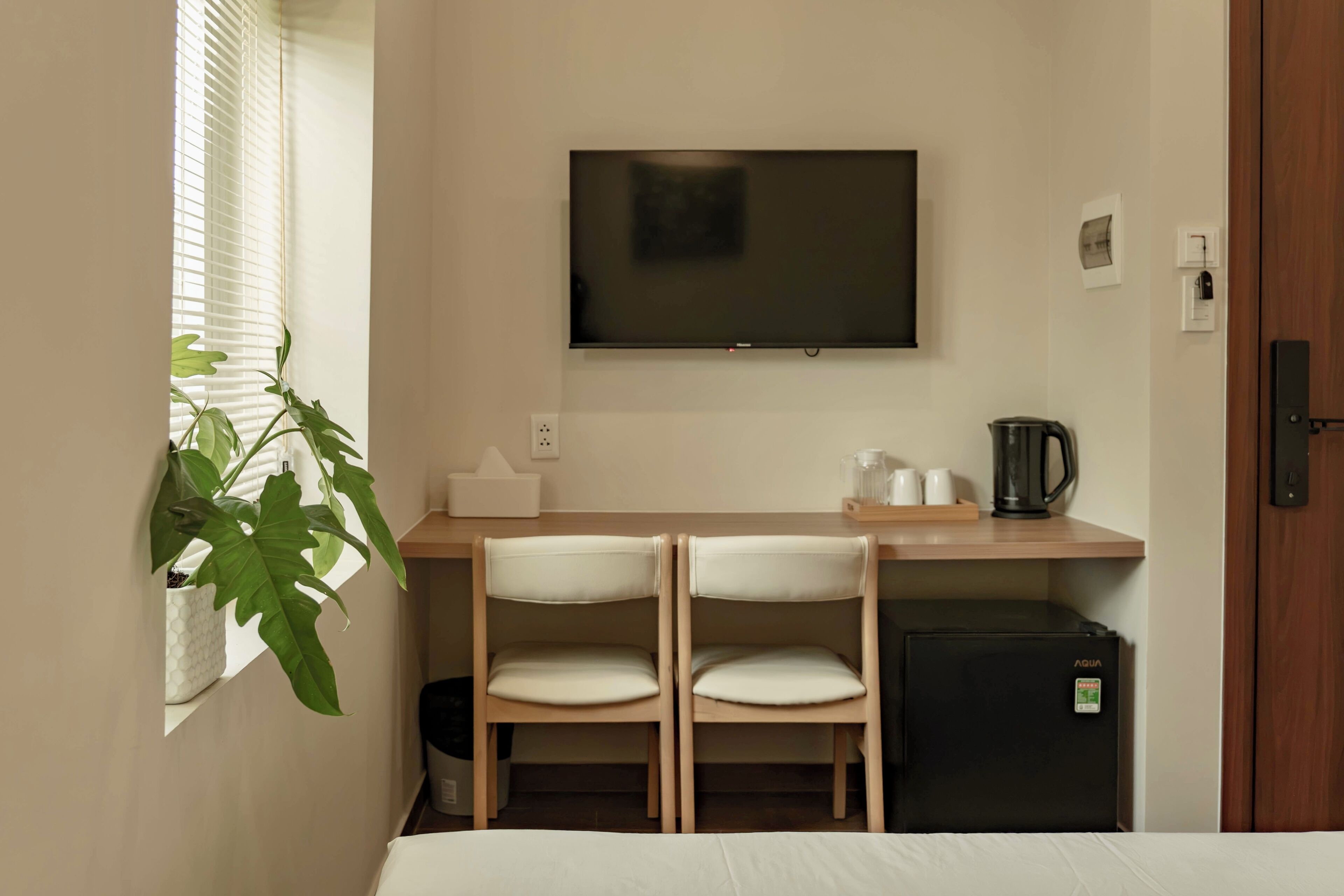 Quarto de hotel compacto com uma cama, uma mesa com duas cadeiras, uma televisão, um refrigerador pequeno e um vaso de planta.