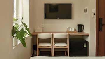 Quarto de hotel compacto com uma cama, uma mesa com duas cadeiras, uma televisão, um refrigerador pequeno e um vaso de planta.
