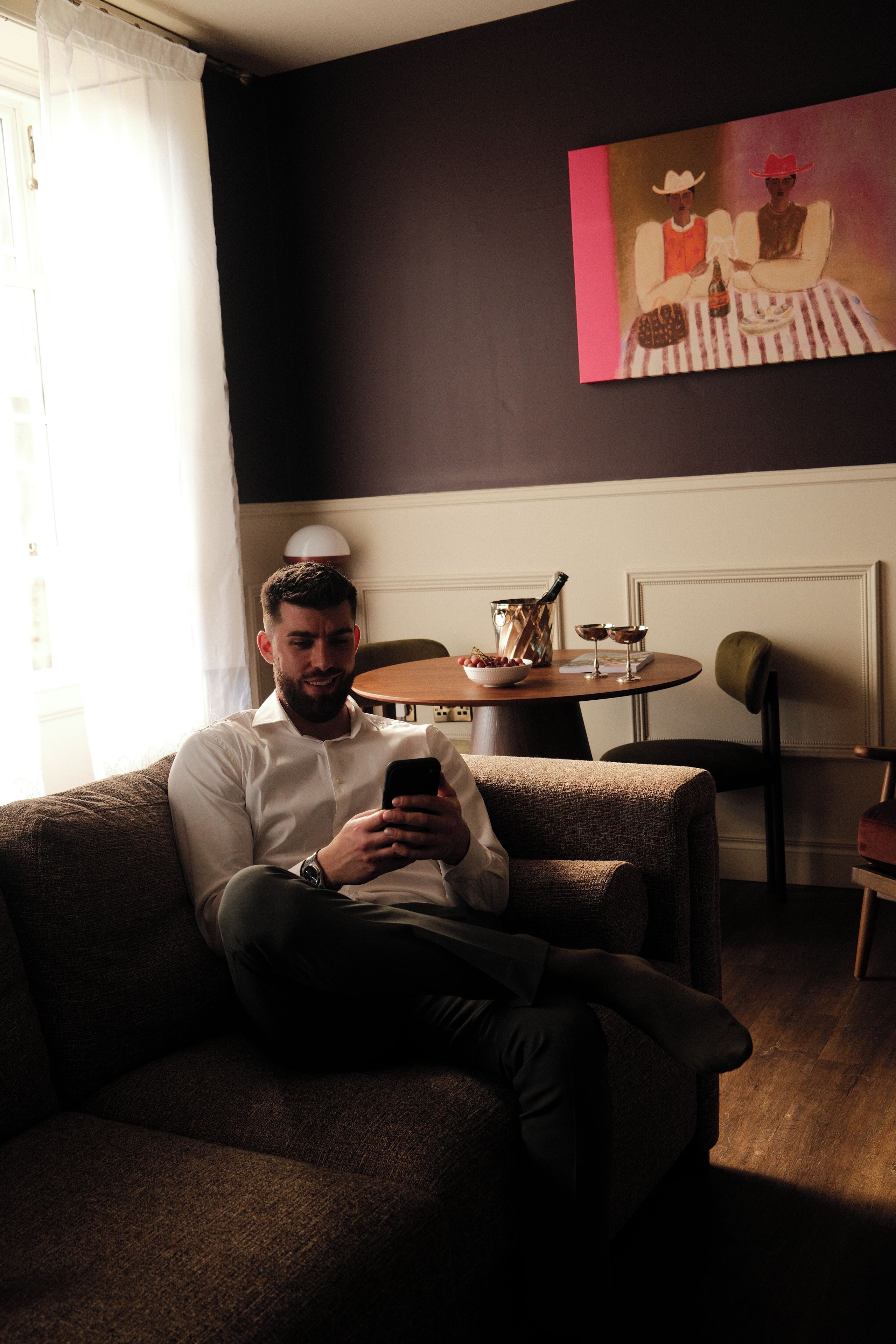 Um homem sentado num sofá usando um smartphone num quarto com um quadro na parede, uma mesa com um jarro e copos, e uma cadeira.