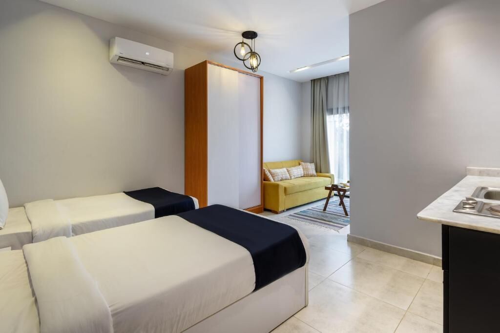 Quarto de hotel com duas camas, um sofá, área de refeições e cozinha compacta.