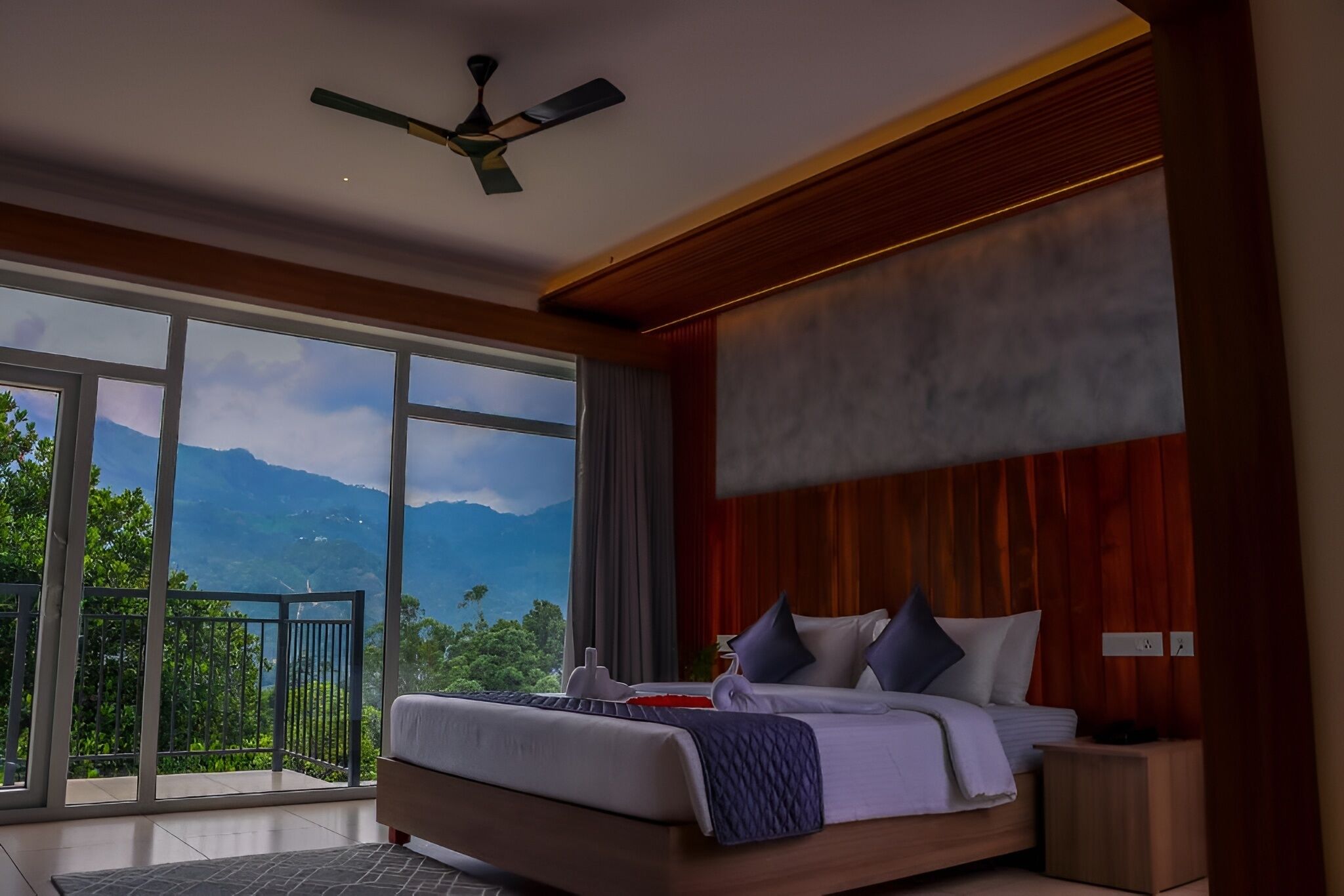 Quarto com uma cama grande, um ventilador de teto, vista para montanhas pela janela e uma varanda.