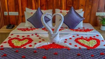Cama com arranjo de toalha em formato de coração e almofadas, rodeada por pétalas de rosa vermelhas.