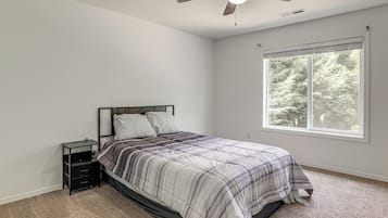 Quarto com cama, mesa de cabeceira, ventoinha no teto e janela com vista para árvores.