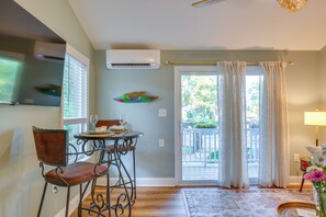 Uma área de refeições com mesa e cadeiras, uma janela com persianas, um aparelho de ar condicionado e vista para o exterior através de uma porta com cortinas.