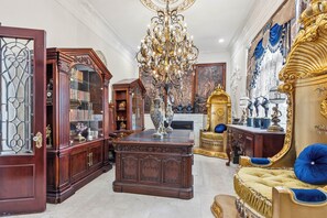 Um quarto opulento com uma grande escrivaninha de madeira, um imponente candelabro e uma poltrona dourada semelhante a um trono.