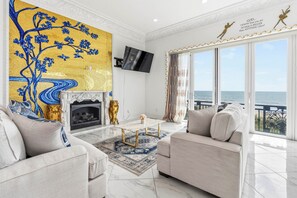 Uma sala de estar com lareira, TV na parede e vista para o mar através de grandes janelas.