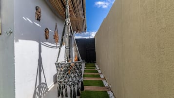 Uma trilha estreita com uma parede branca de um lado e uma parede bege do outro, conduzindo a uma área coberta com um detalhe em macramê pendurado.