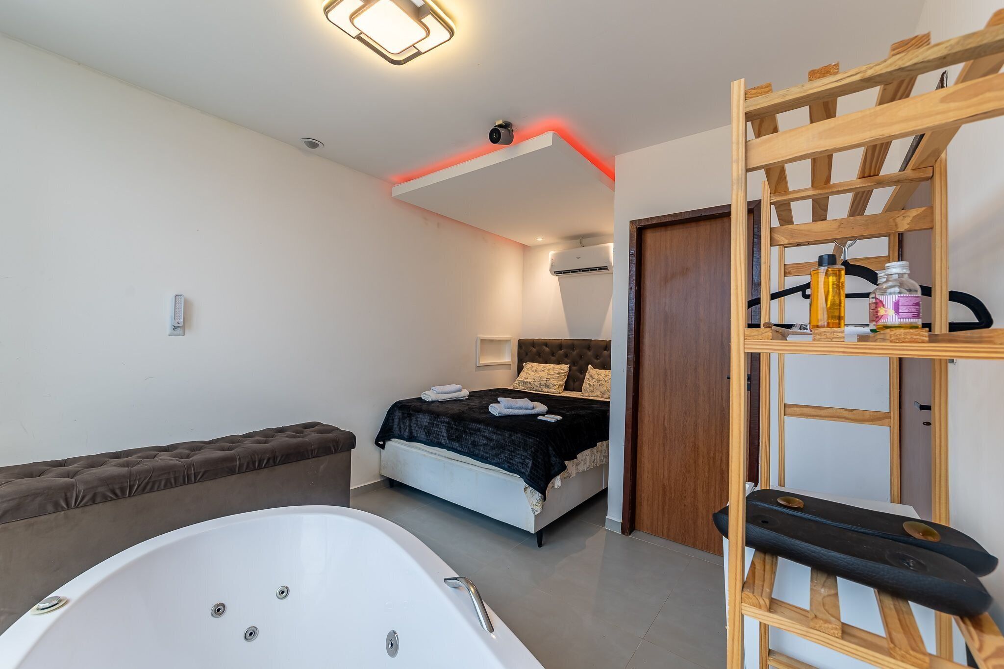 Quarto com cama, prateleira de madeira com artigos de higiene, banheira e ar-condicionado fixado na parede.