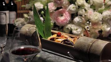 Uma mesa com arranjo floral, um copo de vinho tinto e uma caixa de madeira com alimentos.