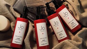 Três frascos vermelhos de loção e condicionador corporais da Jo Loves, um frasco preto com bomba e um sabonete branco sobre uma superfície de tecido bege texturizado.