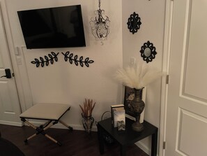 Quarto com televisão de tela plana na parede, lustre decorativo, espelhos fixados na parede, uma pequena mesa com um vaso e livros, e uma cadeira dobrável.