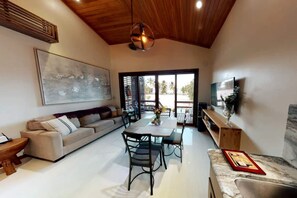 Uma sala de estar ampla com teto alto, um sofá, uma mesa de jantar e uma televisão fixada na parede.