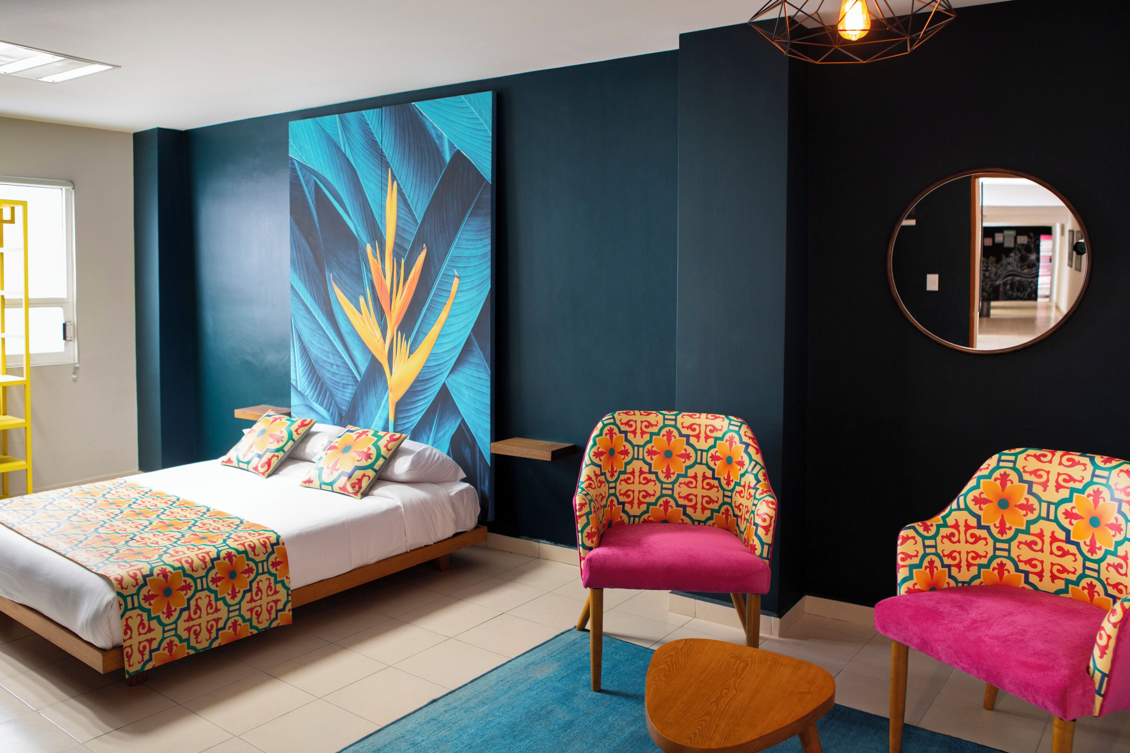 Quarto de hotel com uma cama, duas cadeiras, um grande mural na parede, um espelho e uma janela.