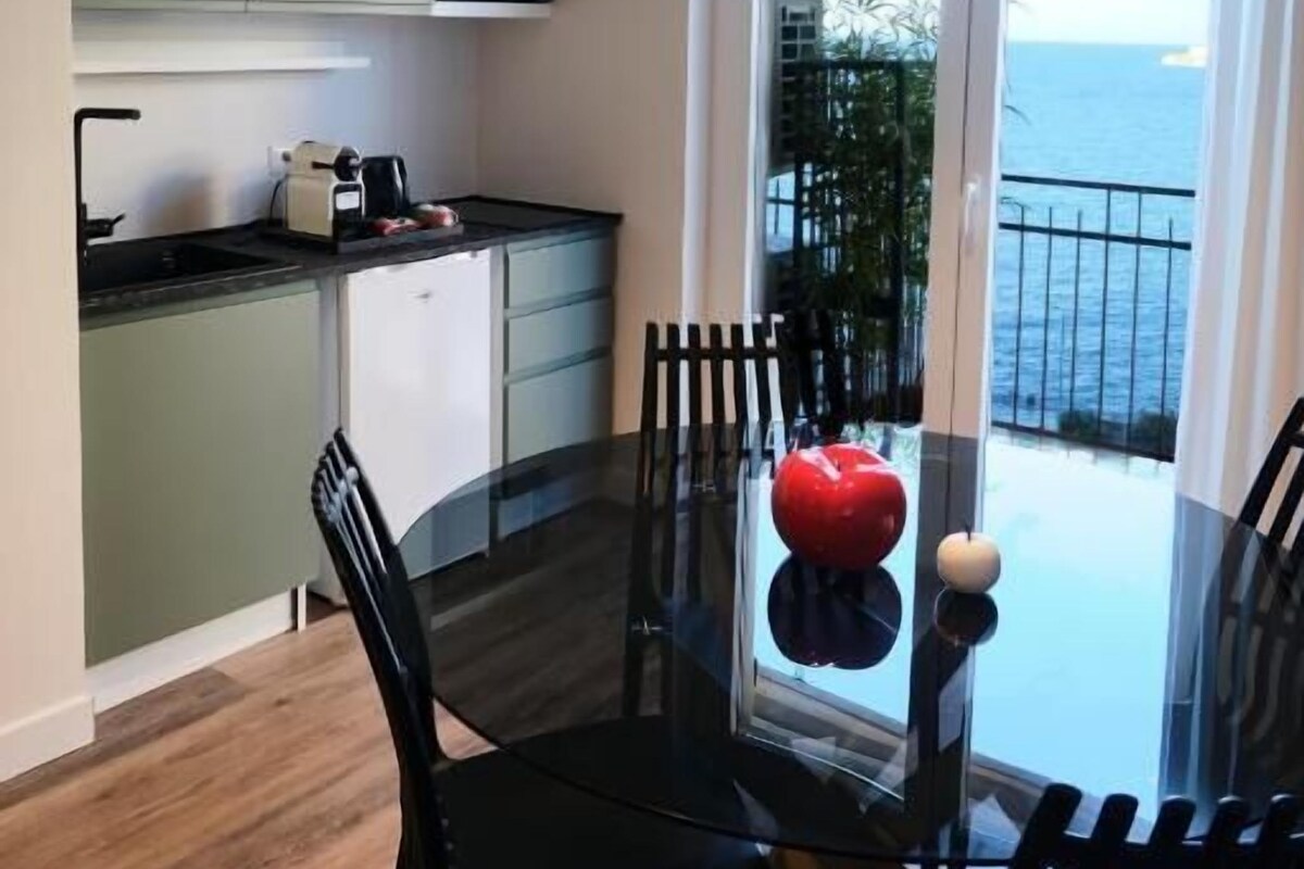 Espaço de refeições com mesa de vidro, cadeiras pretas e vista para o mar através de amplas janelas.