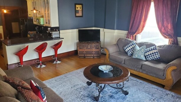 Uma sala de estar com um sofá, uma mesa de centro e uma televisão. Há cadeiras vermelhas e um armário ao fundo.
