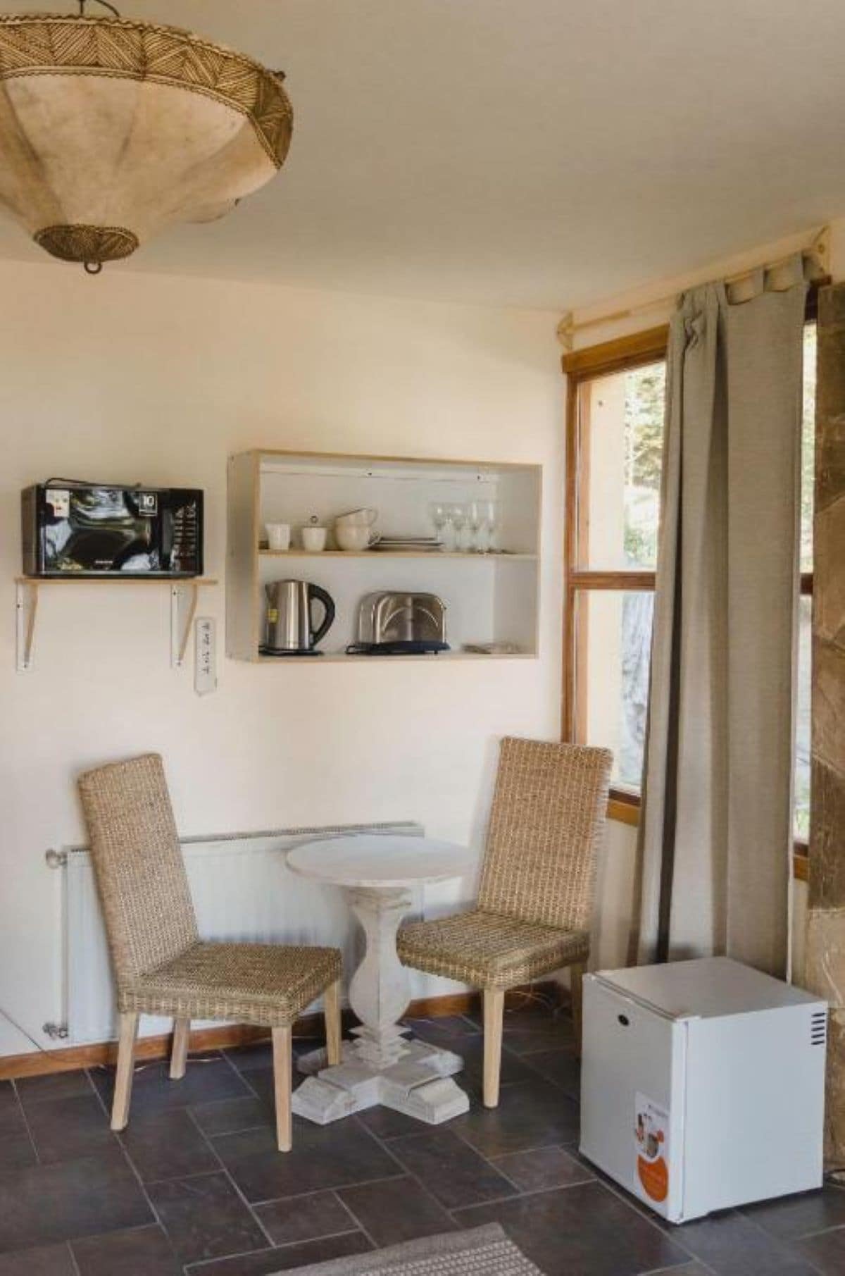 Quarto com mesa de jantar, duas cadeiras de vime, geladeira branca, prateleira com utensílios de cozinha e luminária pendente.