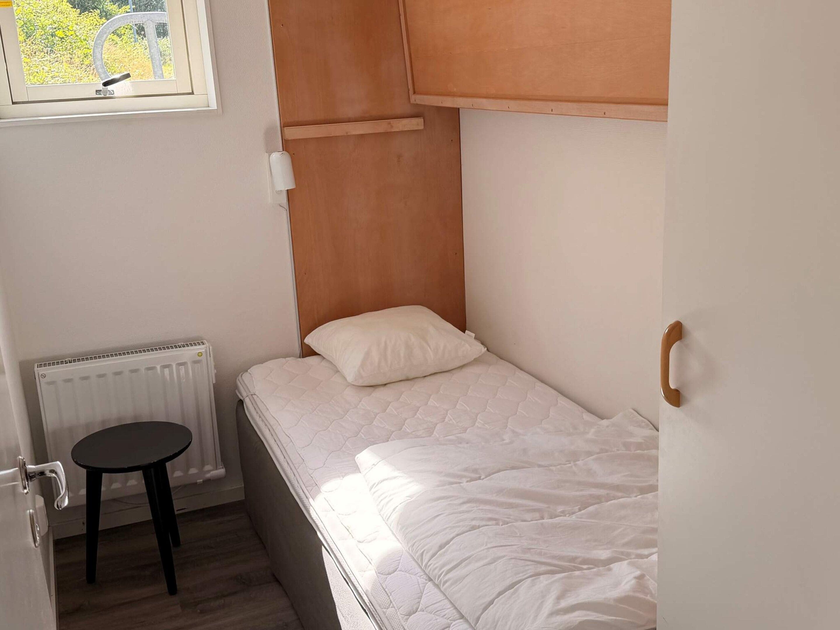 Quarto pequeno com uma cama de solteiro, cabeceira de madeira, uma mesinha e um radiador.
