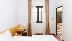 Quarto de hotel com uma cama, uma escrivaninha e uma cadeira. Há uma janela com cortinas e uma porta que dá para uma área externa.