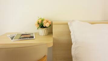 Quarto de hotel com cama, mesa de cabeceira, uma cadeira e uma pequena mesa com uma revista e um vaso de flores.