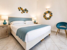 Cama bem arrumada com manta azul, duas mesas de cabeceira com luminárias, uma cadeira azul e arte decorativa na parede.