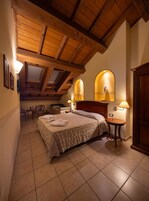 Quarto com teto de madeira, uma cama com cabeceira, duas mesas de cabeceira com abajures, uma cadeira e uma pequena mesa com um abajur.