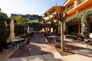 Uma área de café ao ar livre com mesas e cadeiras, cercada por vegetação e um edifício com telhado em azulejos.
