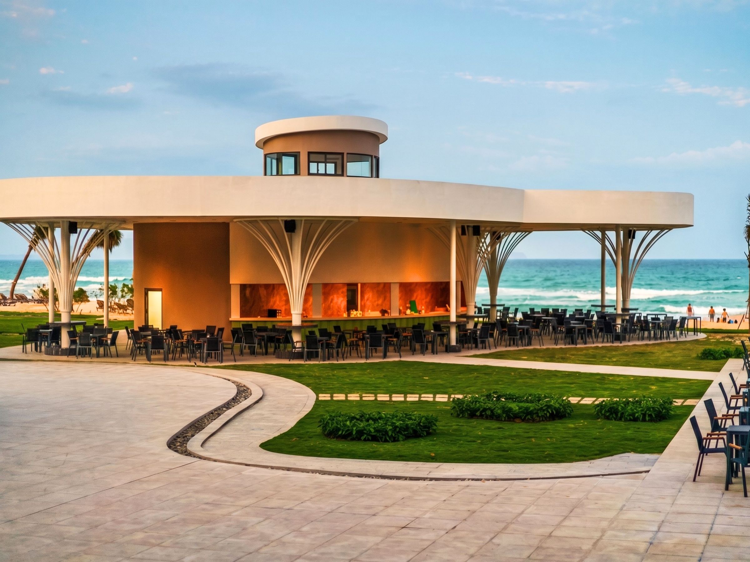 Um edifício moderno com uma seção central circular, rodeado por uma área de estar externa coberta, com cadeiras e mesas, com vista para uma praia.