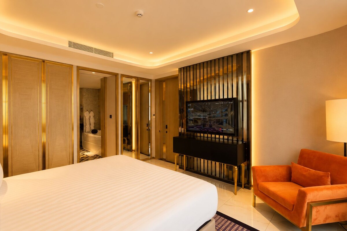 Quarto de hotel com uma cama grande, TV na parede, área de estar com sofá e banheiro visível por uma porta aberta.