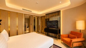 Quarto de hotel com uma cama grande, TV na parede, área de estar com sofá e banheiro visível por uma porta aberta.