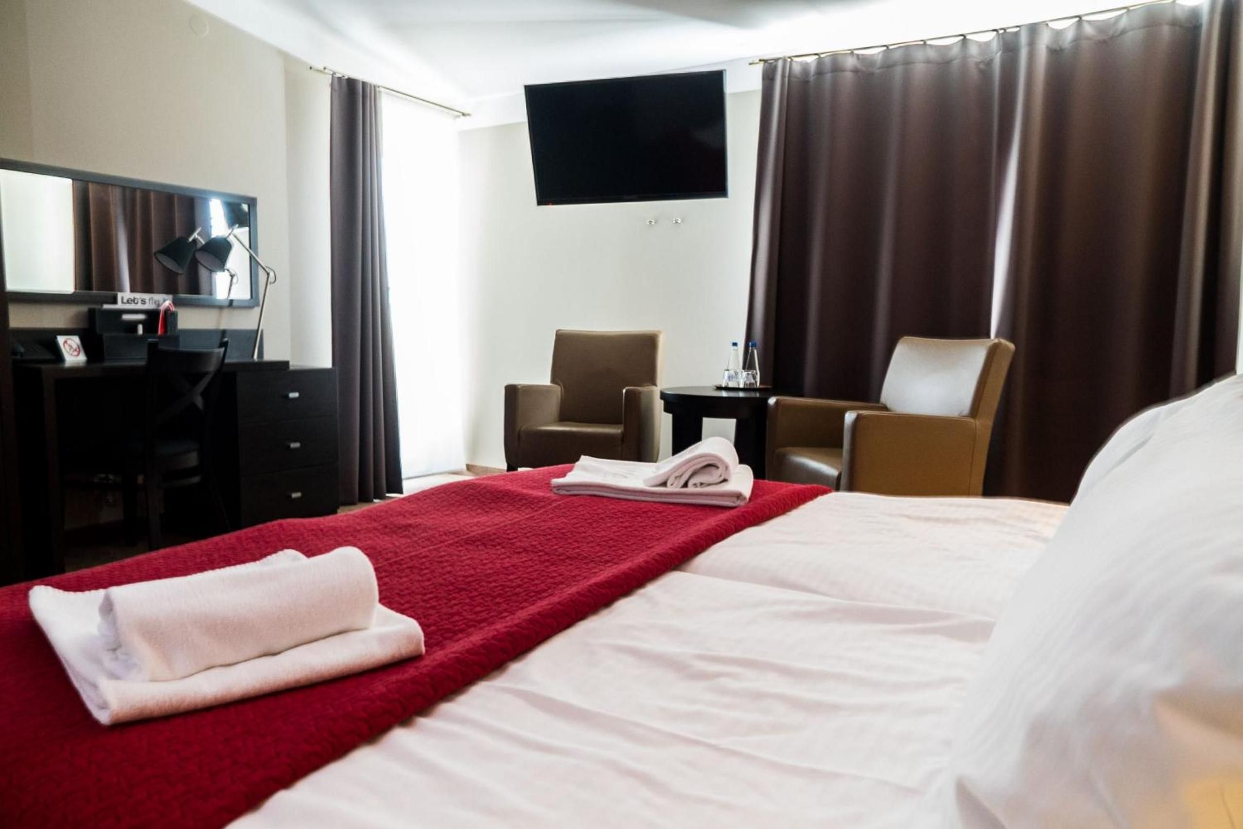 Quarto de hotel com cama, escrivaninha, cadeira, televisão e janela com cortinas.