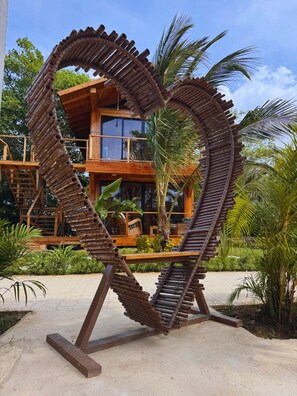 Escultura em formato de coração feita com tubos de bambu em um ambiente externo, com um edifício de madeira ao fundo.