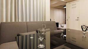 Quarto de hotel com sofá, mesa de vidro com taças de vinho e uma porta com espelho refletindo o quarto.