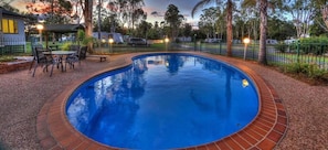 Piscina com borda de tijolos, cercada por uma grade e área de estar ao ar livre.