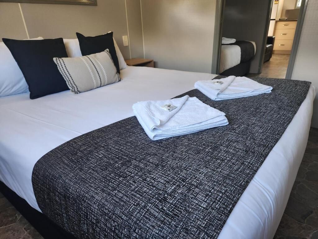Uma cama bem arrumada com lençóis brancos e um cobertor cinza texturizado, uma pequena mesa de cabeceira e uma cadeira em um quarto de hotel.