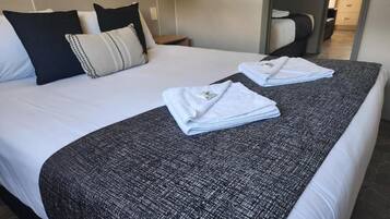 Uma cama bem arrumada com lençóis brancos e um cobertor cinza texturizado, uma pequena mesa de cabeceira e uma cadeira em um quarto de hotel.