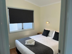 Quarto de hotel com cama, janela com cortina, mesa de cabeceira e luz acoplada na parede.