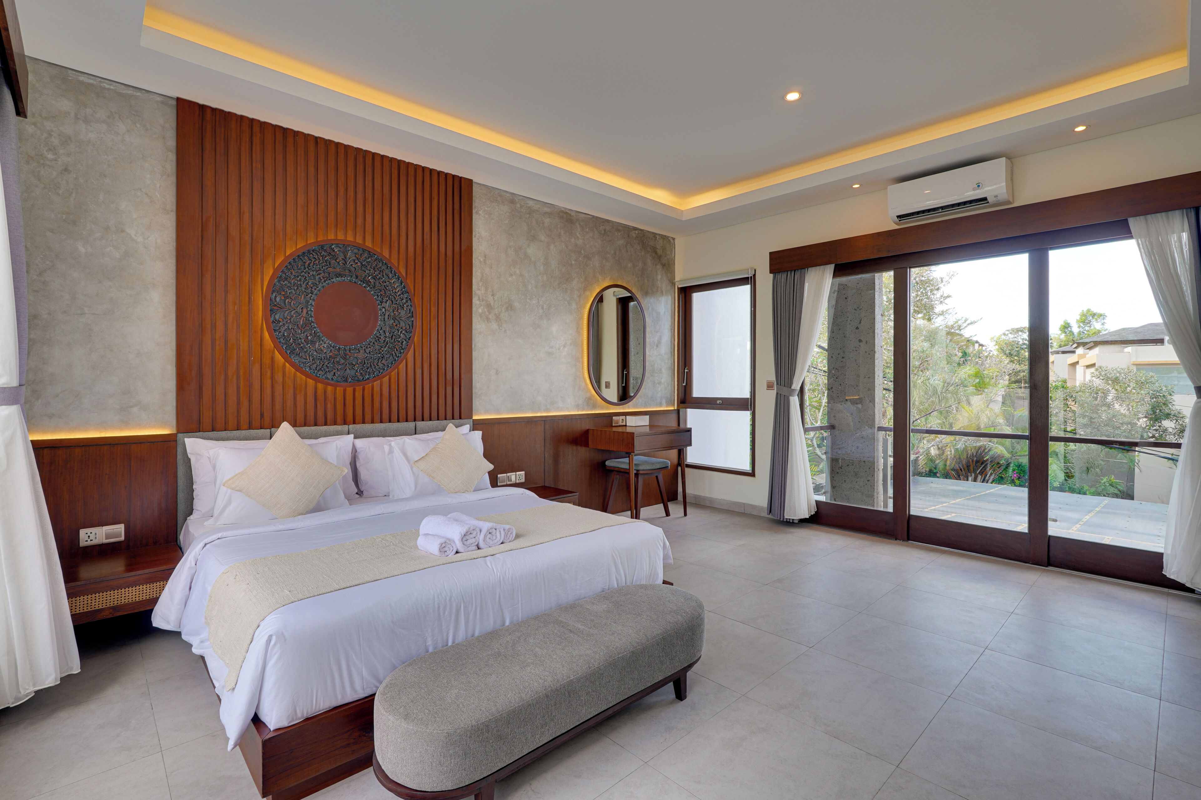 Quarto de hotel moderno com uma cama grande, uma mesa de trabalho, um banco, um detalhe decorativo circular na parede e uma janela ampla com vista.